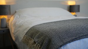 Uma cama com travesseiro branco e manta decorada. Existem duas luminárias de cabeceira, uma de cada lado da cama.