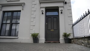 Um edifício com porta escura, duas plantas em vasos, um muro de pedra e uma cerca de metal preto.