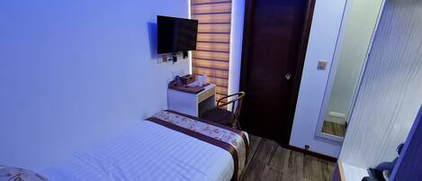 Quarto compacto com cama, mesa de cabeceira pequena, cadeira, guarda-roupa e televisão na parede.