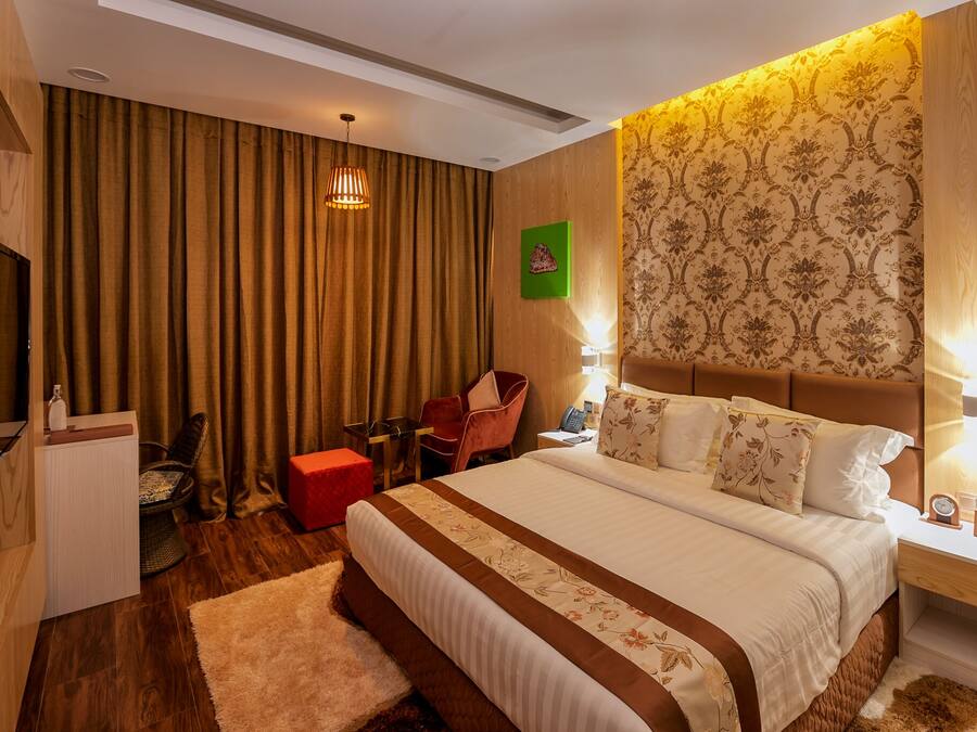 Quarto de hotel com uma cama grande, televisão, escrivaninha, cadeira e um banquinho vermelho.