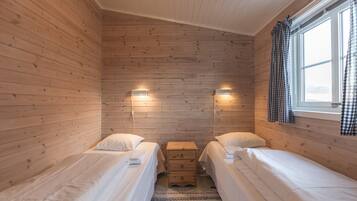 Quarto com paredes de madeira e duas camas individuais com roupa de cama branca. Há duas luminárias fixadas na parede e uma janela com cortinas xadrez.