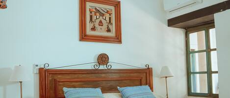 Um quarto com cama de madeira, duas mesas de cabeceira, luminária pendente, ar-condicionado fixado na parede e janela com persianas verdes.