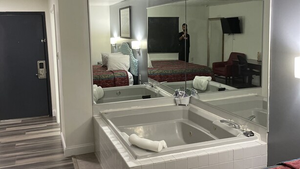 Quarto de hotel com um espelho grande, uma banheira, uma cama com colcha vermelha estampada e uma porta.