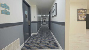 Corredor de hotel com piso carpetado, sinalização de área de máquinas e um quadro na parede.