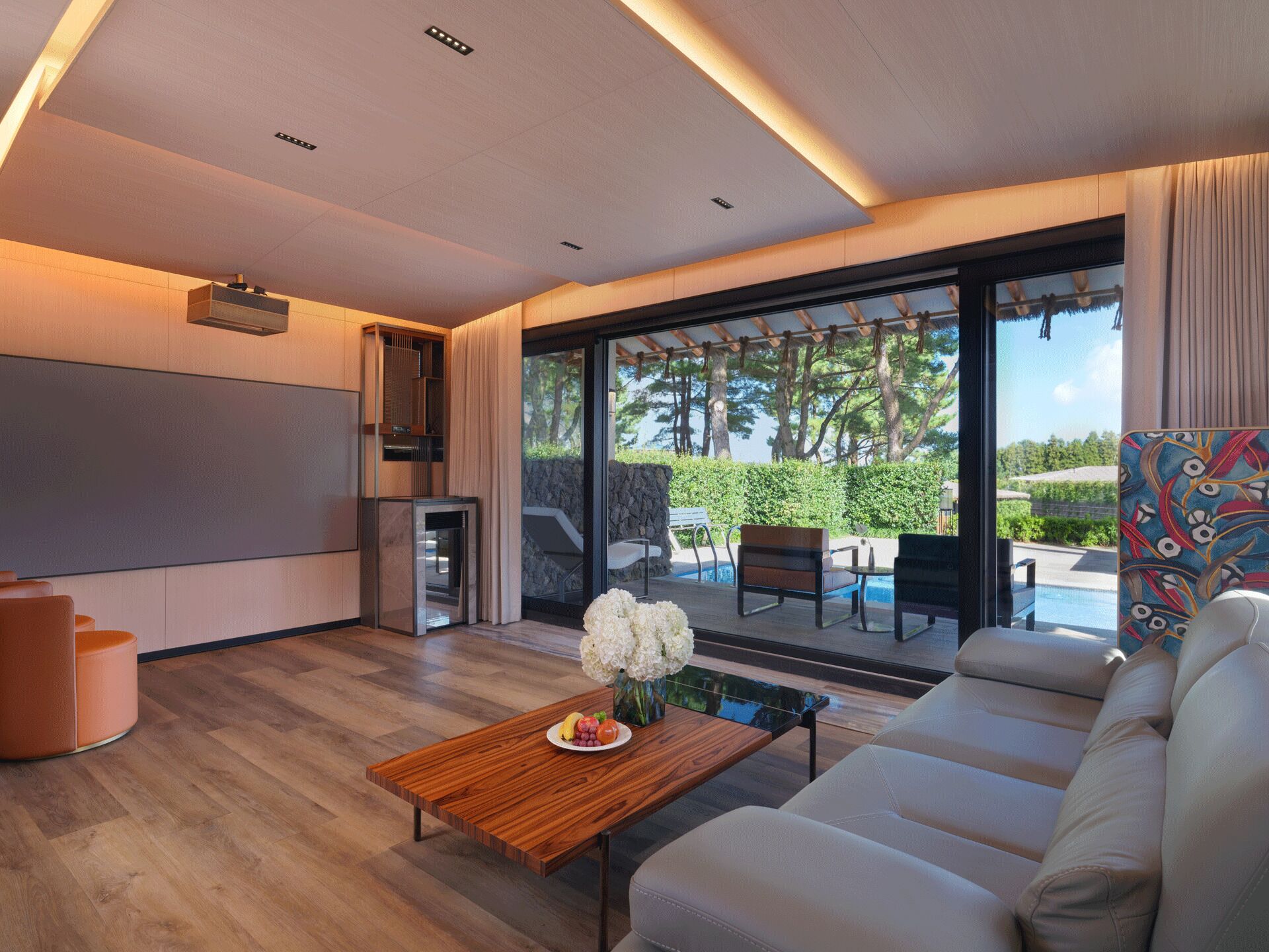 Uma sala de estar moderna com um sofá, uma mesa de centro com frutas e vista para uma piscina e vegetação.