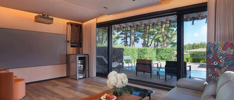 Uma sala de estar moderna com um sofá, uma mesa de centro com frutas e vista para uma piscina e vegetação.