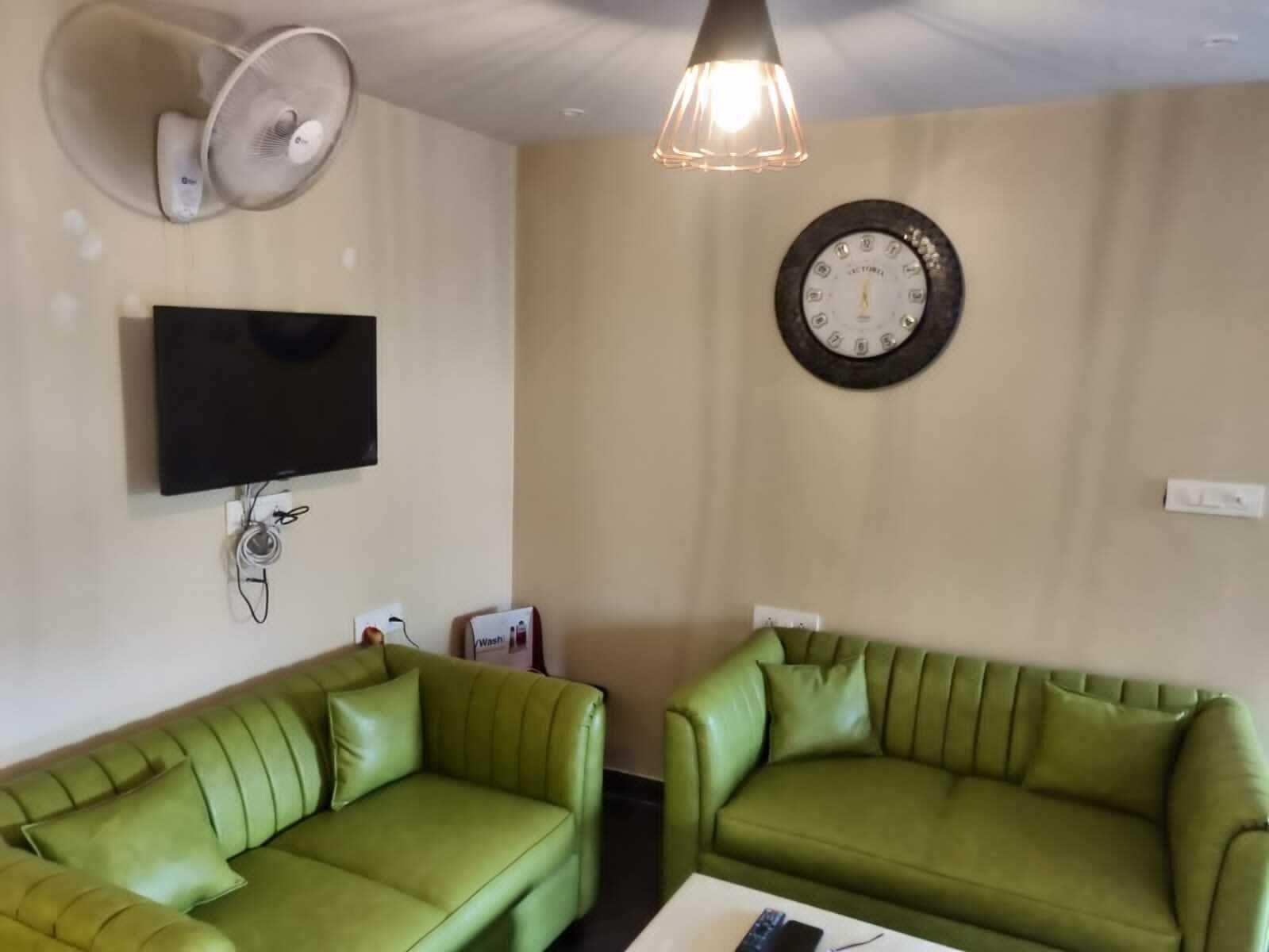 Um quarto com dois sofás de couro verde, uma televisão de tela plana, um relógio de parede e um ventilador de teto.