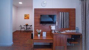 Quarto de hotel com secretária de madeira, televisão de tela plana na parede, relógio, mesa pequena com cadeiras e quadro na parede.