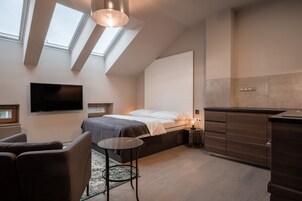 Quarto moderno com cama, televisão, mesinha e sofá. Apresenta claraboias e uma área de cozinha visível.