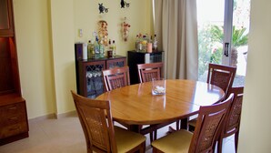 Uma área de refeições com uma mesa de madeira redonda e seis cadeiras, um armário com diversos frascos, e decorações nas paredes.