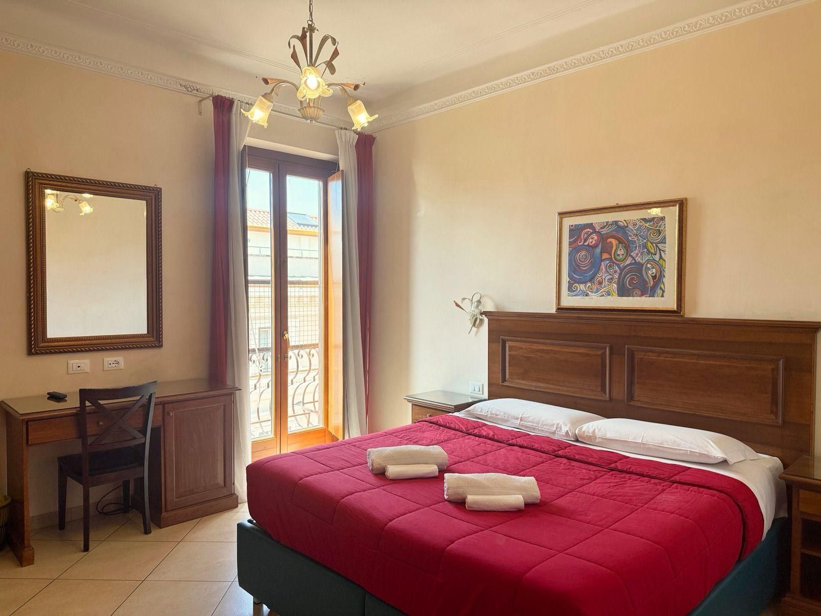 Quarto de hotel com cama, escrivaninha, cadeira, espelho, quadro na parede e janela com cortinas.