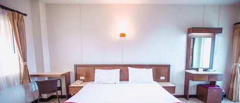 Quarto de hotel com uma cama grande, uma escrivaninha, uma cadeira, uma mesa de cabeceira, um espelho e uma janela com cortinas.