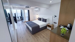 Quarto de hotel moderno com uma cama grande, mesa de cabeceira e pequena cozinha.