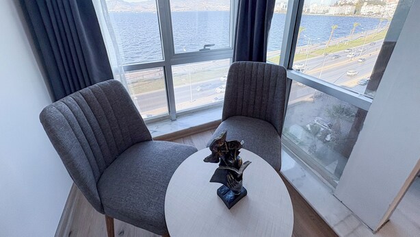 Quarto com uma grande janela com vista para um corpo de água e paisagem urbana. Há duas poltronas cinzas e uma pequena mesa branca com uma escultura.