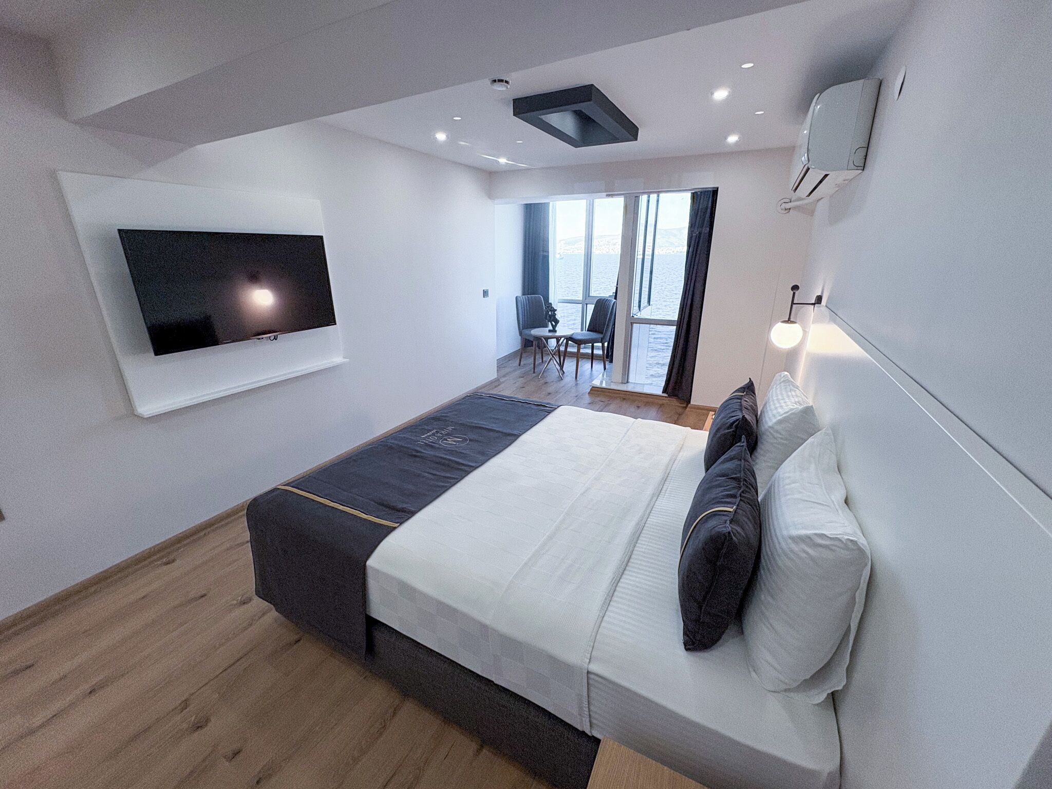 Quarto de hotel moderno com uma cama grande, uma televisão de tela plana na parede e vista para o mar pela janela.