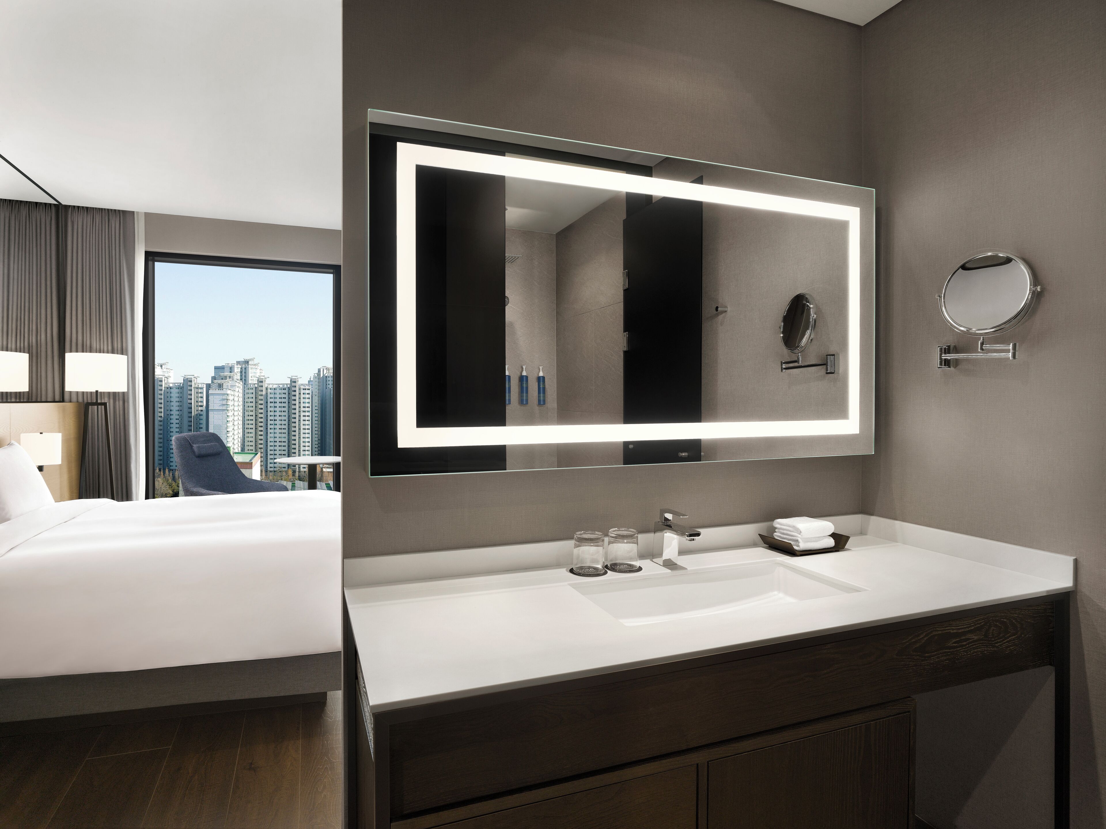Quarto de hotel moderno com uma cama grande, banheiro com toucador, um espelho amplo e vista para o horizonte da cidade.
