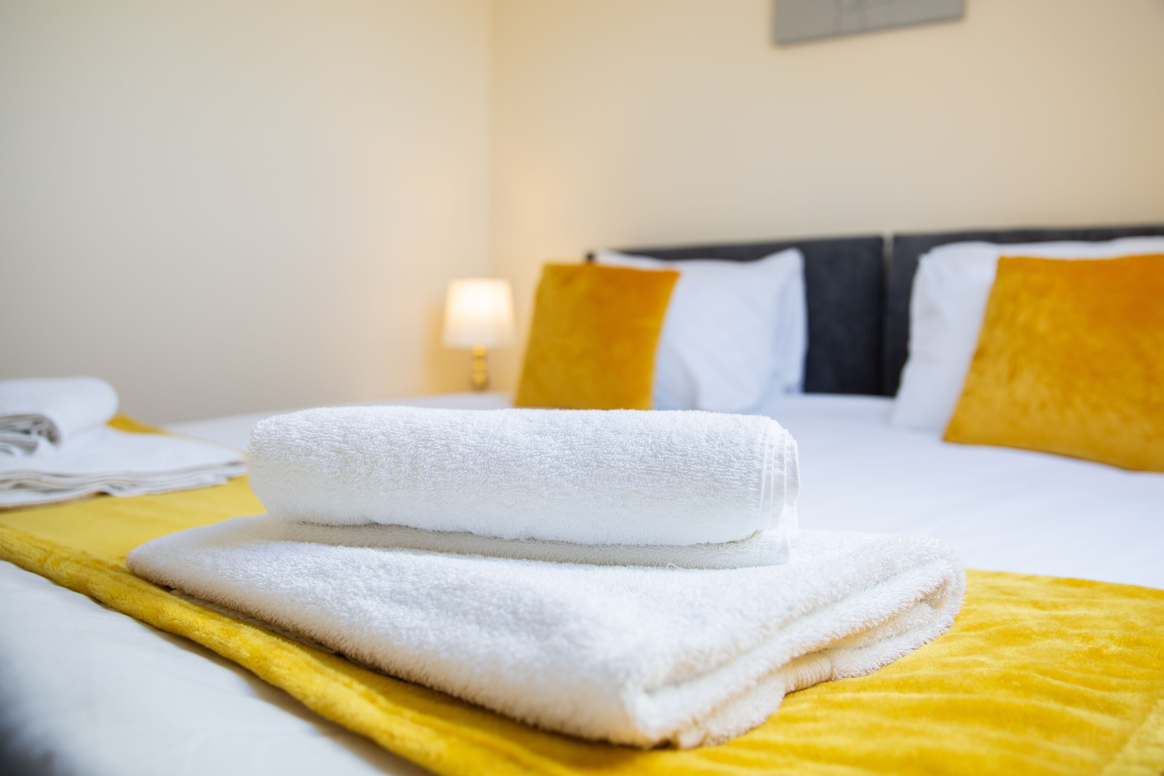 Cama bem arrumada com roupa de cama branca e amarela, uma toalha branca dobrada e uma luminária de cabeceira.
