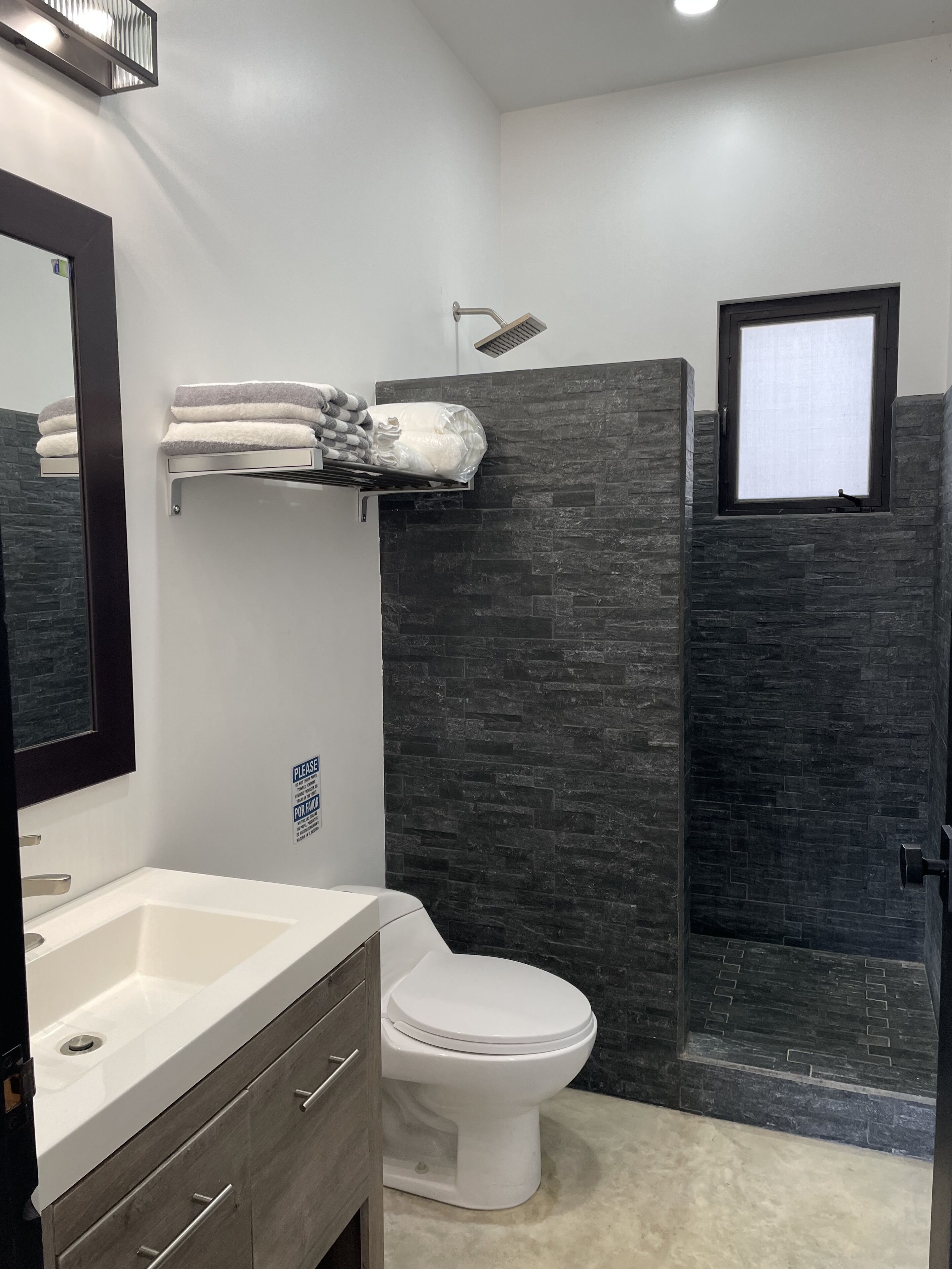 Uma casa de banho moderna com uma sanita branca, uma sanita e uma cabine de duche com parede revestida a mosaicos escuros, além de uma janela.