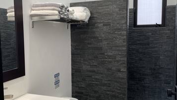 Uma casa de banho moderna com uma sanita branca, uma sanita e uma cabine de duche com parede revestida a mosaicos escuros, além de uma janela.