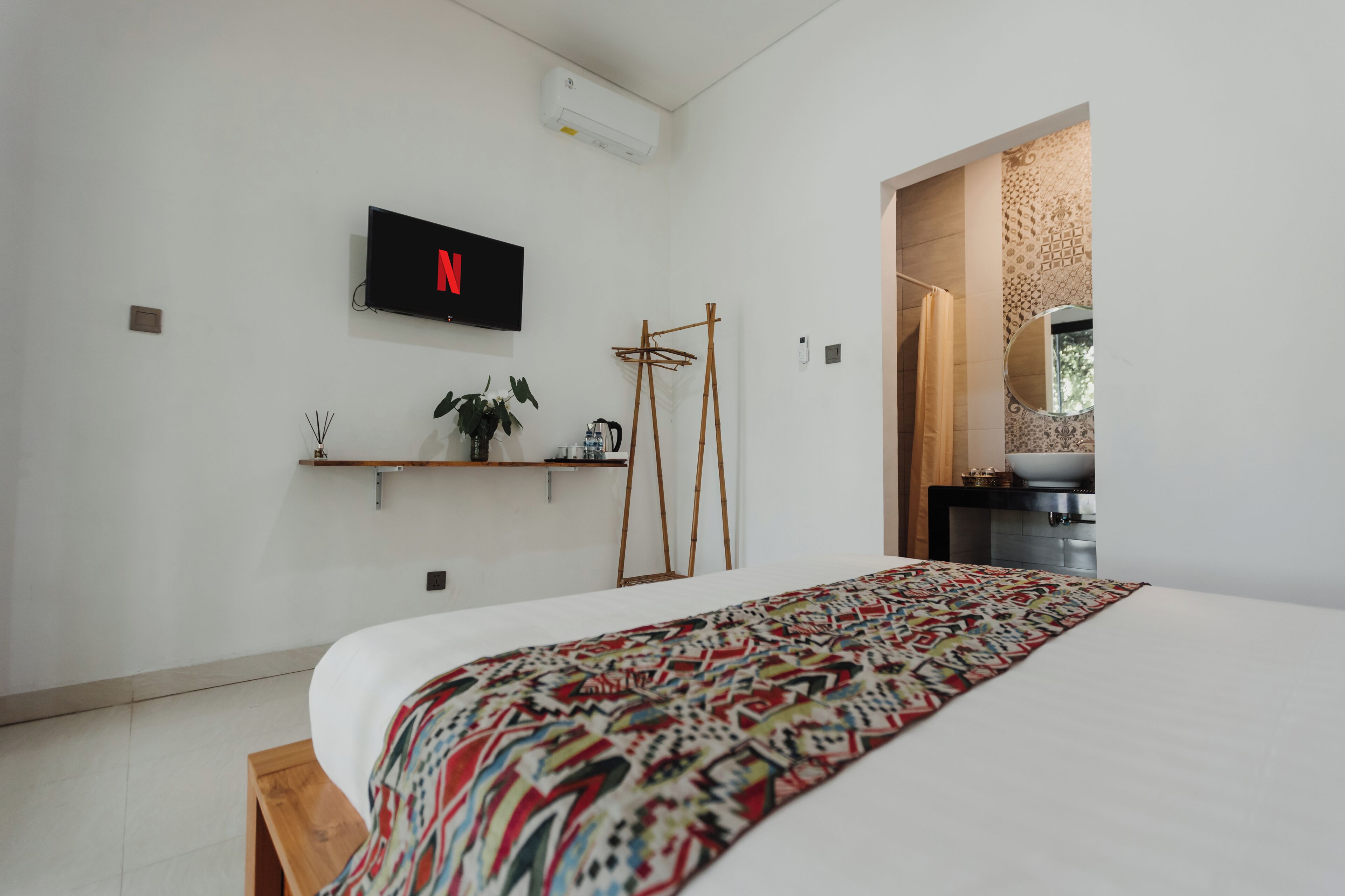 Quarto com cama, televisão fixada na parede exibindo o logotipo da Netflix, prateleira com planta e varal de madeira.