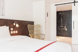 Quarto de hotel moderno com cama, cadeira de vime, área de chuveiro e cabeceira de madeira.