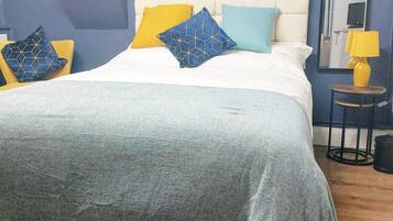 Uma cama bem arrumada com cabeceira branca, manta cinza e várias almofadas em amarelo e azul. Há uma mesa pequena com abajur amarelo ao lado da cama.