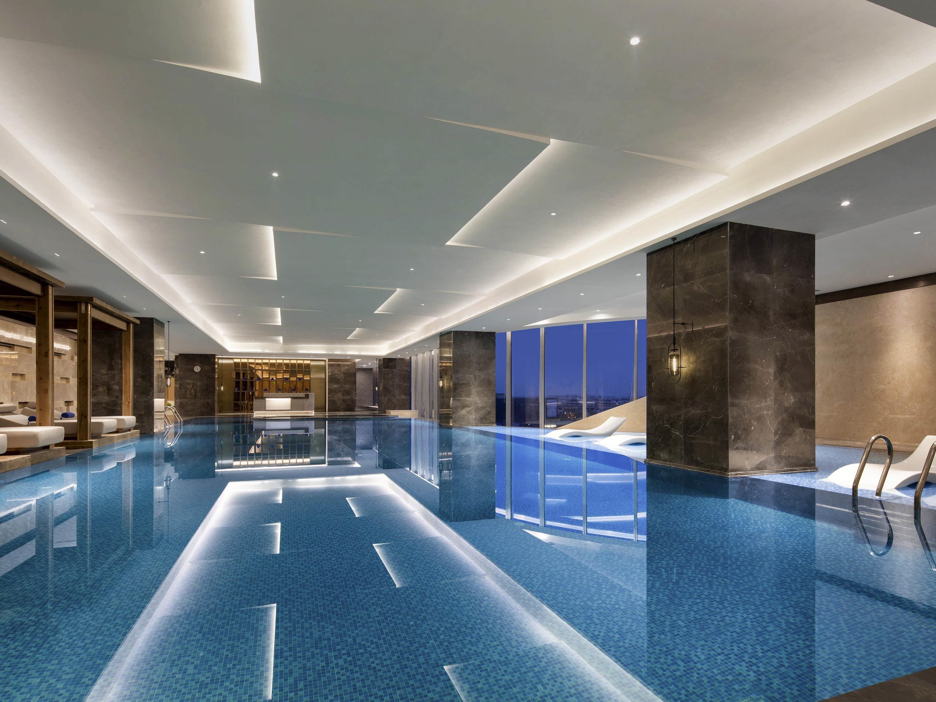 Piscina interior moderna com água cristalina, área de recepção e vista da cidade à noite.