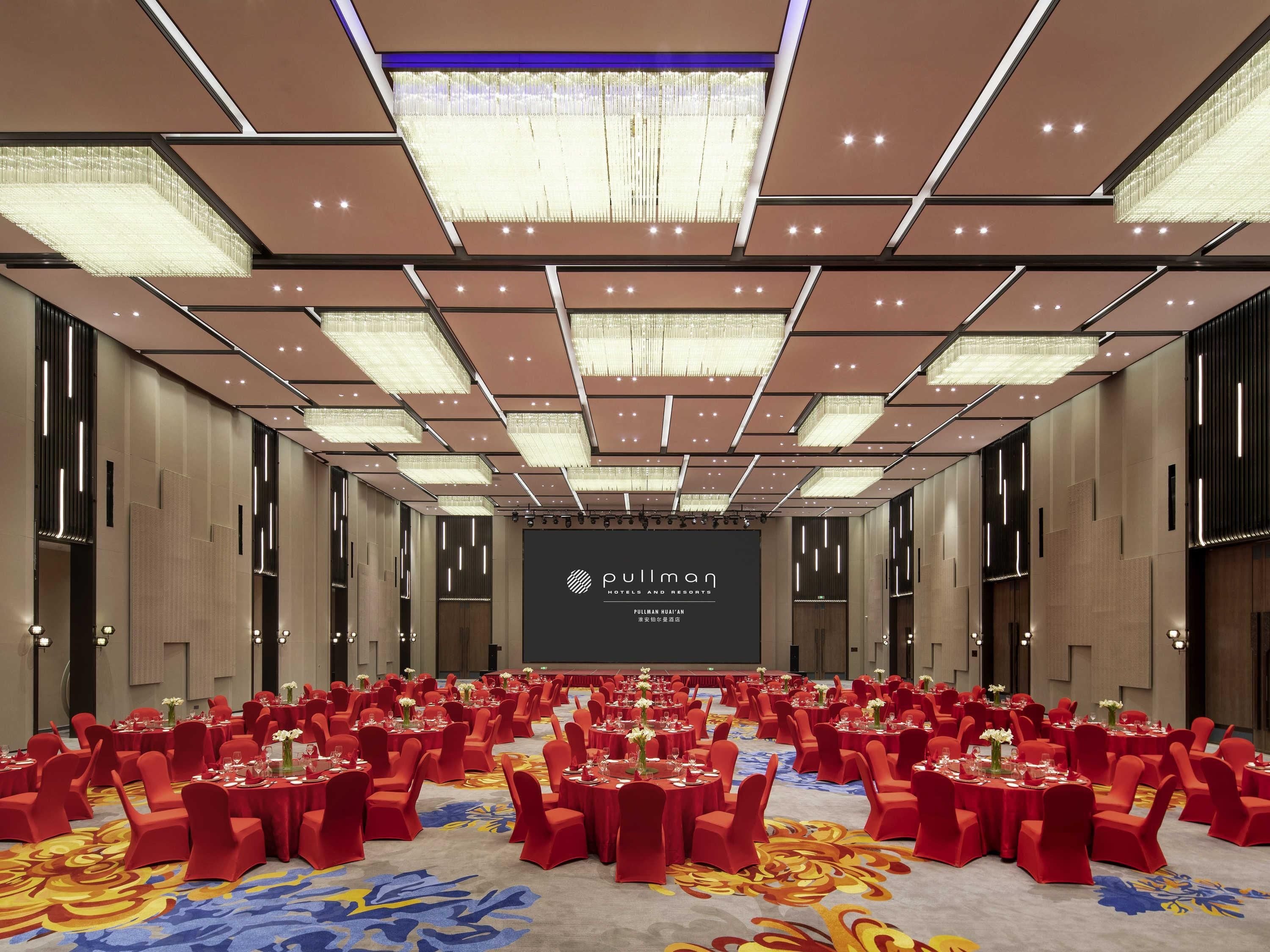 Uma ampla sala de conferências com mesas preparadas para um evento formal, uma tela grande exibindo o nome do hotel e um lustre moderno.