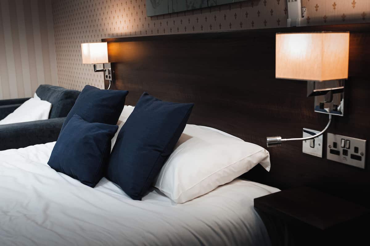 Cama bem arrumada com travesseiros brancos e azul escuro, cabeceira de madeira e duas luminárias fixadas na parede.