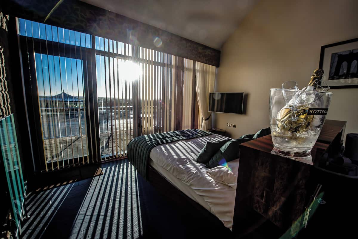 Quarto de hotel com cama, uma janela grande com persianas, minibar e vista para a cidade.