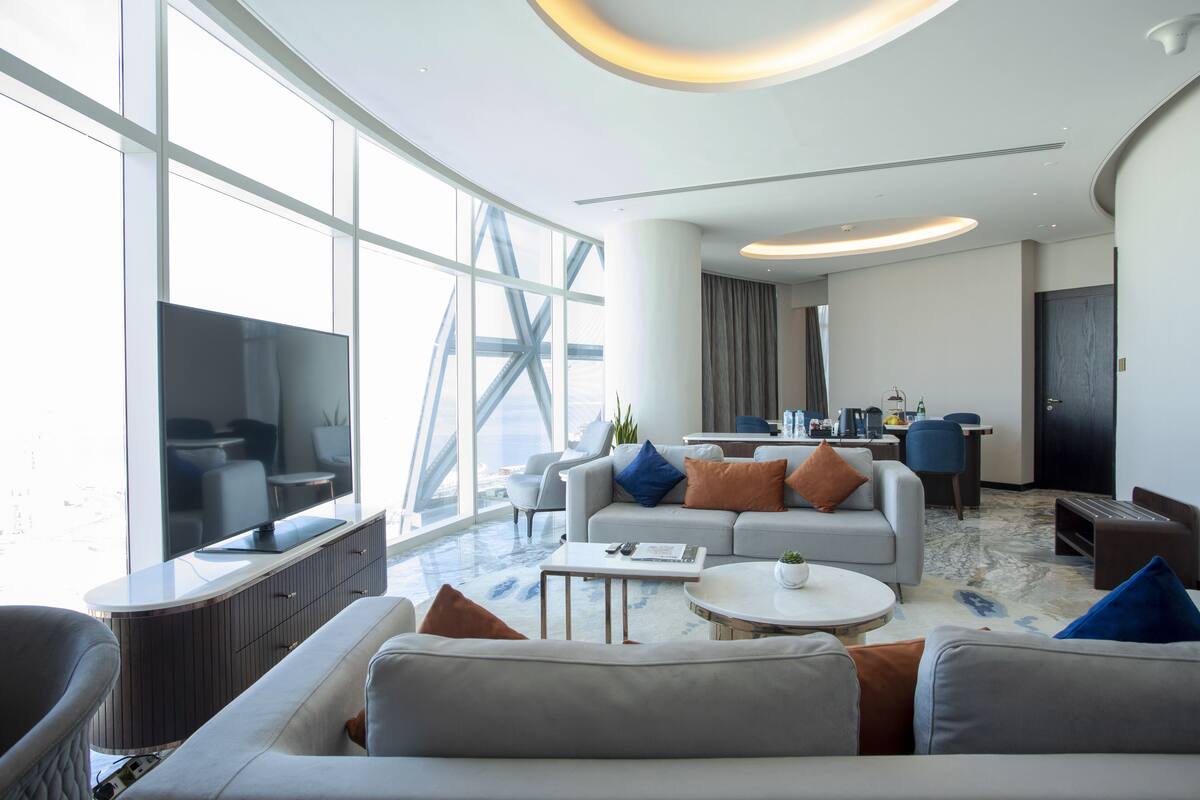 Quarto de hotel moderno com televisão grande, assentos confortáveis, área de refeições e grandes janelas com vista para a cidade.
