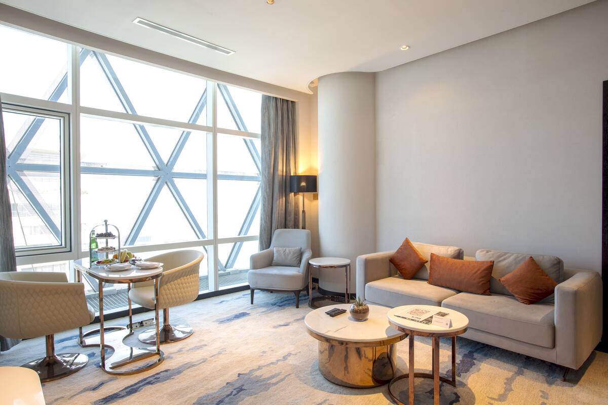 Quarto de hotel moderno com uma janela ampla, um sofá, duas poltronas, uma mesa de centro e uma pequena mesa lateral.