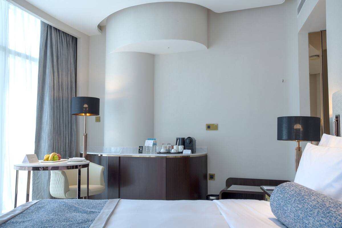 Quarto de hotel com cama, escrivaninha com luminária, cadeira, minibar e uma janela grande com cortinas.