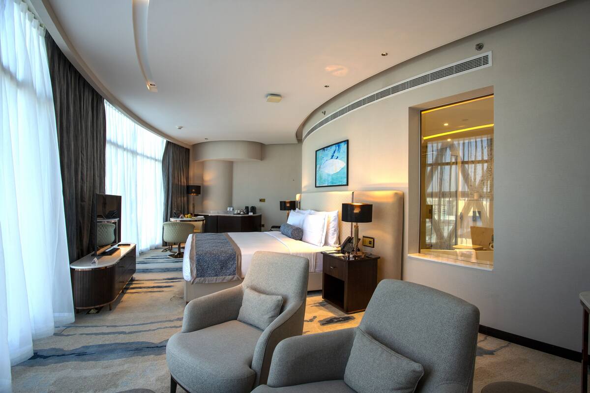 Quarto de hotel com cama, duas poltronas, uma escrivaninha, televisão e uma janela grande.