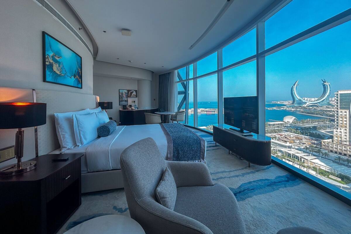 Quarto de hotel moderno com uma cama grande, uma mesa, uma cadeira e uma TV. O quarto possui janelas amplas que oferecem vista para a cidade e uma ponte.
