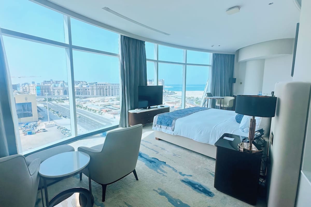 Quarto de hotel com uma janela grande, uma cama, uma escrivaninha, uma cadeira e uma televisão.