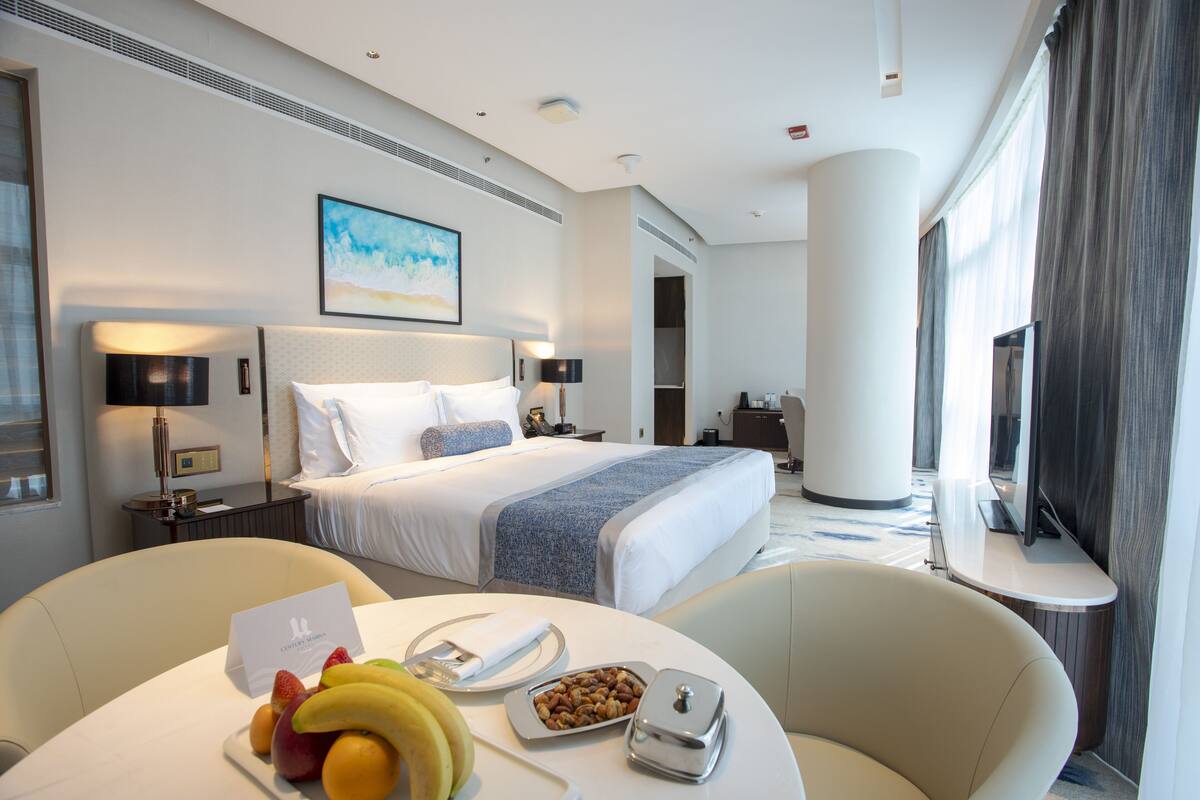 Quarto de hotel moderno com uma cama bem arrumada, uma mesa de jantar com frutas e uma bandeja de nozes, uma TV e uma grande janela com cortinas.