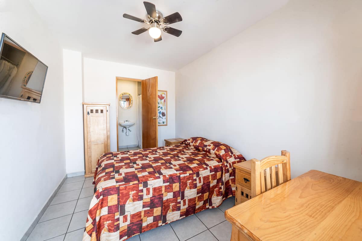 Quarto com cama, mesa de madeira, ventilador de teto, TV fixada na parede e porta que dá para um banheiro.