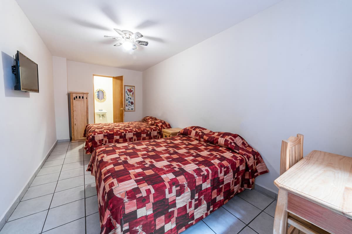 Quarto com duas camas, uma mesa de madeira, um ventilador de teto e uma televisão na parede.