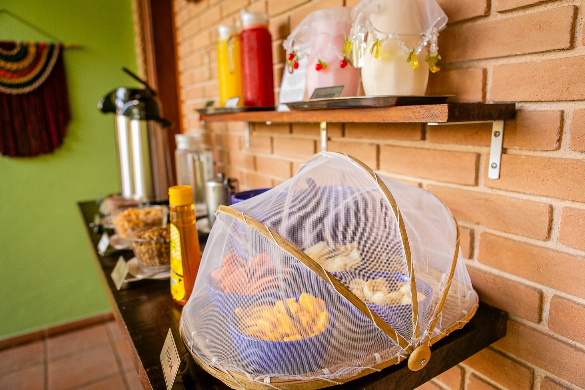 Um balcão de café da manhã com uma cesta de frutas, uma cafeteira e diversos acompanhamentos.