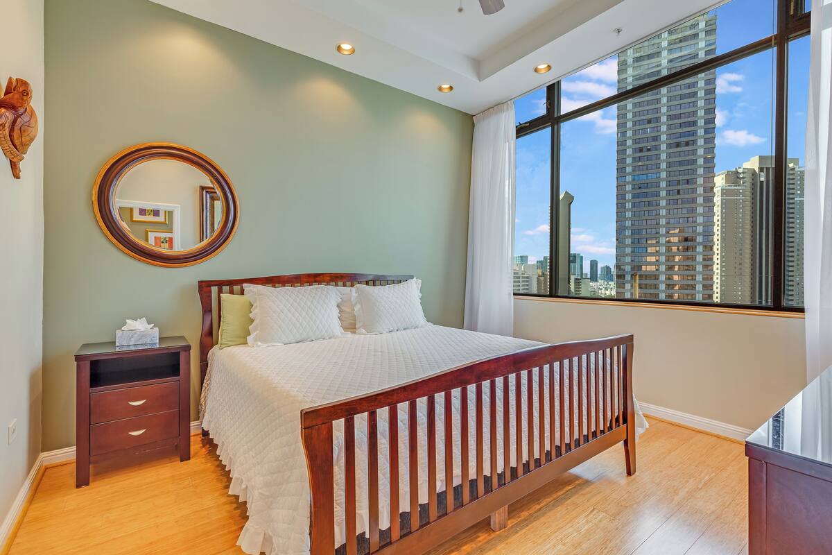 Quarto com cama de madeira, mesa de cabeceira, espelho redondo e grande janela com vista para a cidade.