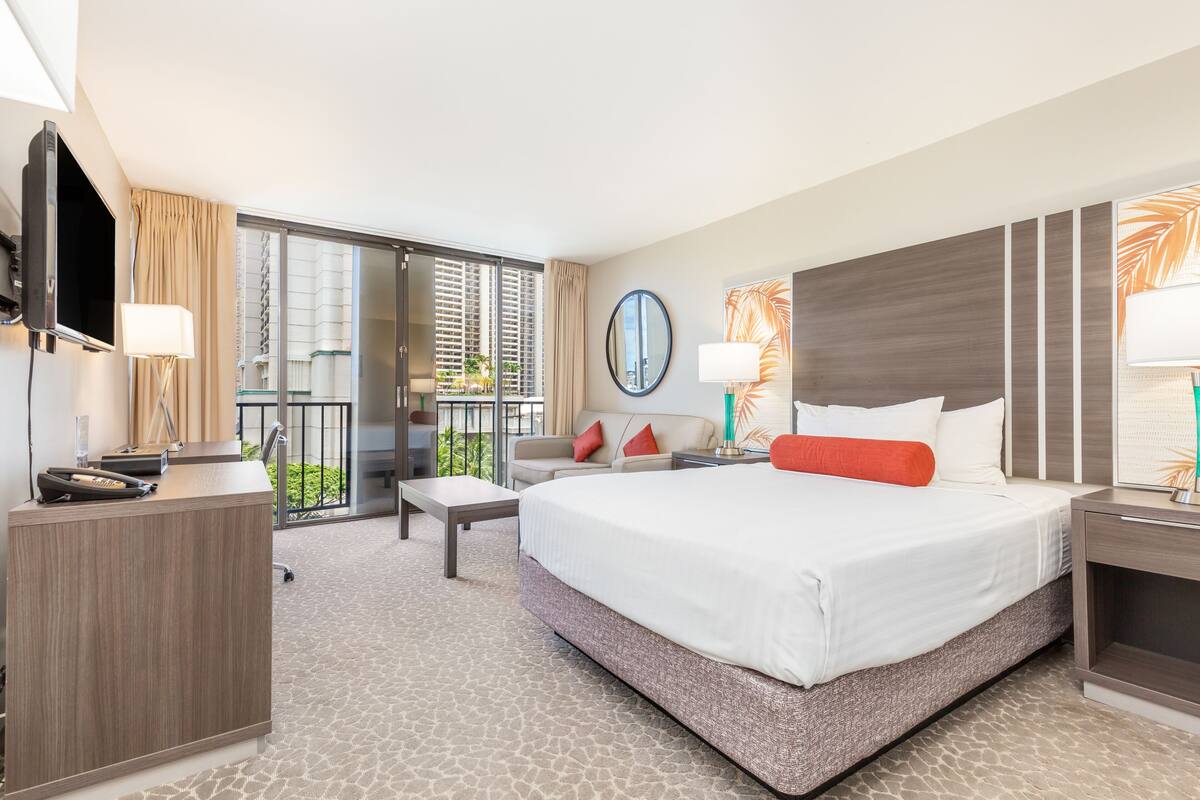 Quarto de hotel com uma cama grande, uma escrivaninha, televisão e varanda com vista para edifícios.