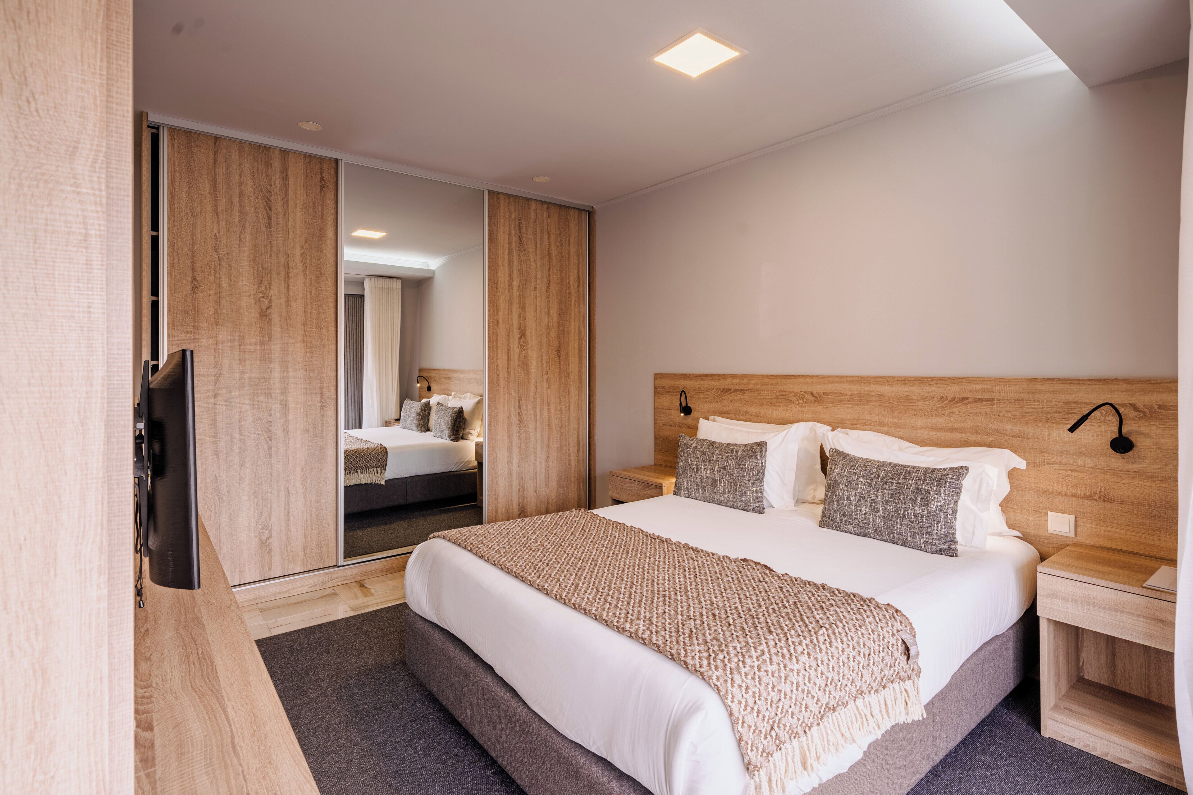 Quarto de hotel moderno com uma cama grande, cabeceira de madeira, mesas de cabeceira, um espelho e um guarda-roupa.