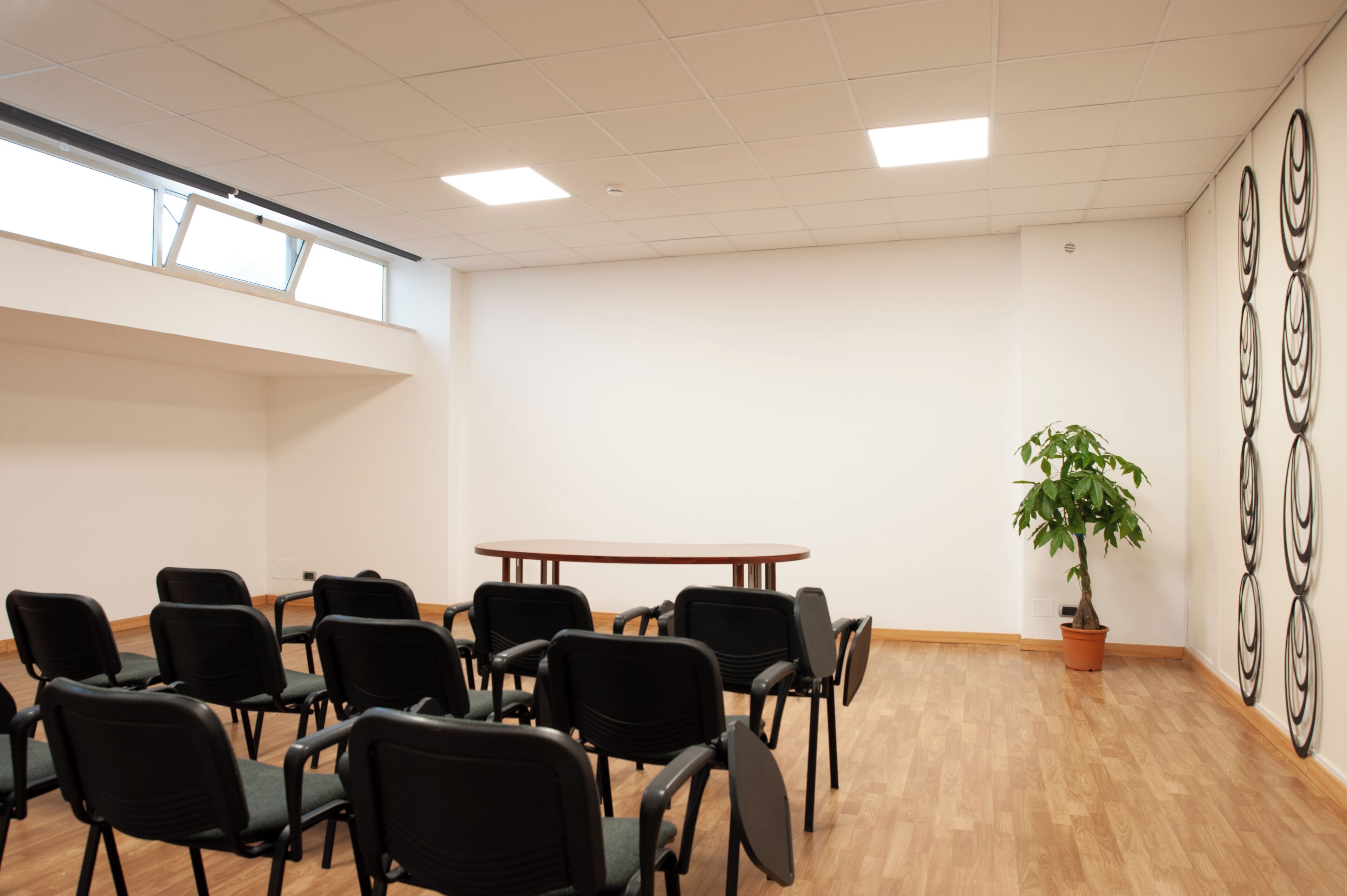 Uma sala de conferências com uma mesa redonda, cadeiras pretas, um vaso de planta e um painel decorativo.