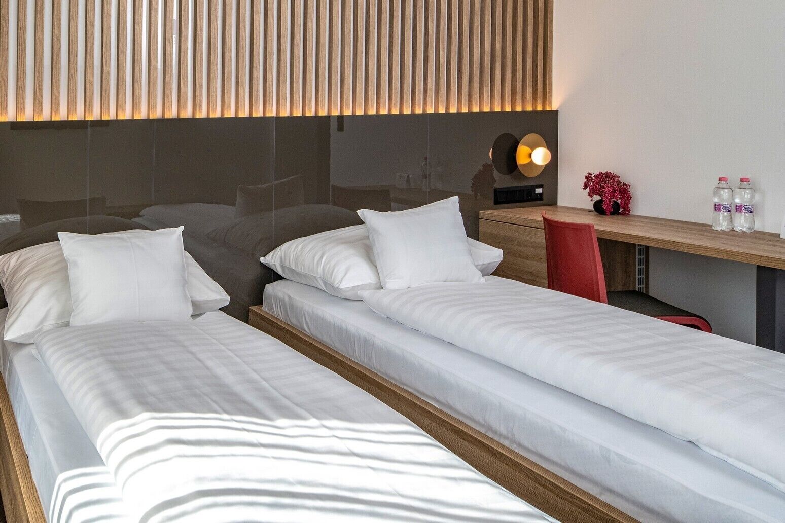 Quarto de hotel com duas camas de solteiro, cabeceira de madeira, mesa de cabeceira com abajur e uma pequena cadeira vermelha.
