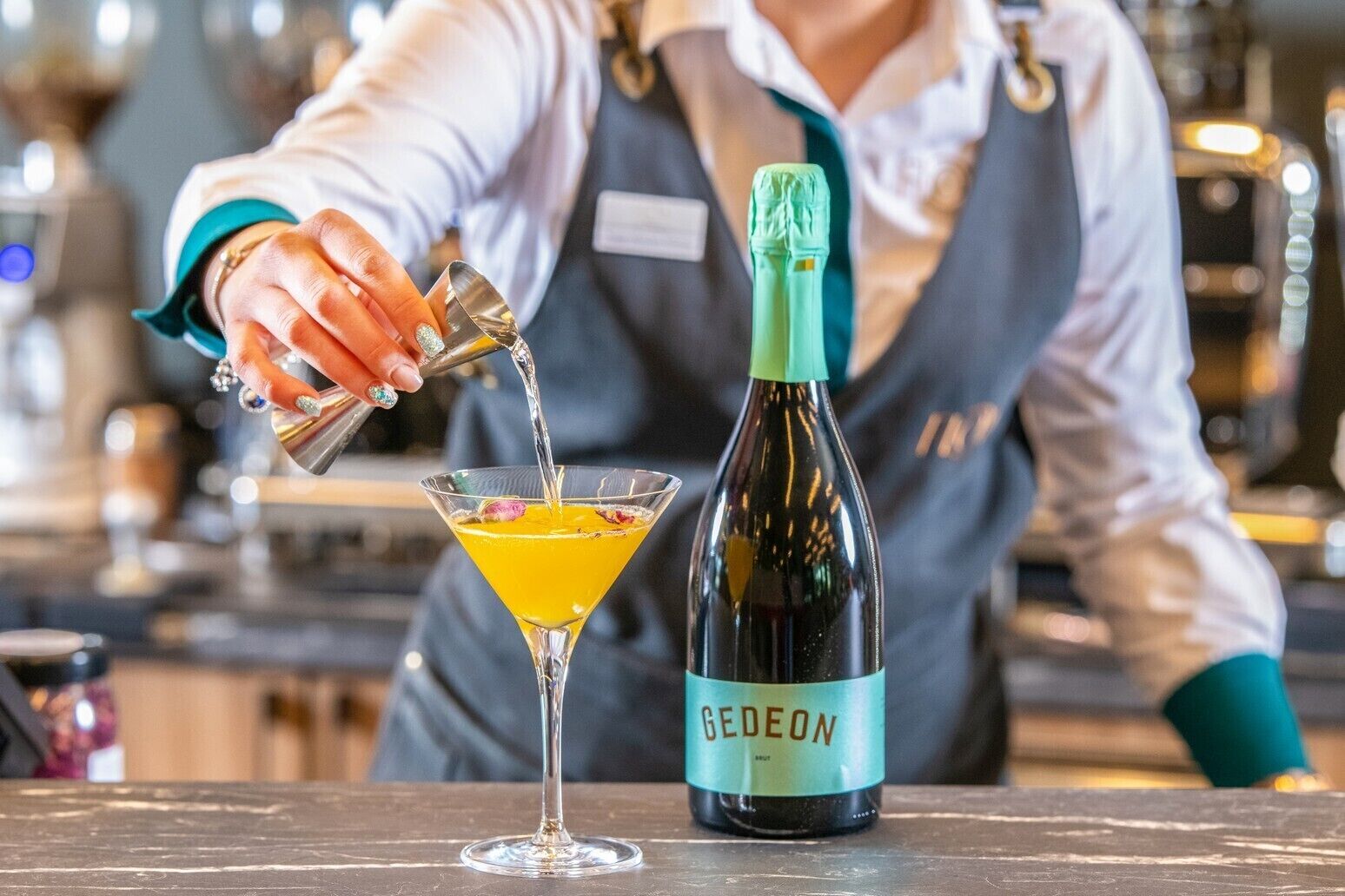 Um bartender servindo uma bebida em uma taça de martini ao lado de uma garrafa com a etiqueta 'Gedeon'.