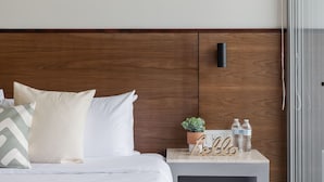 Quarto moderno com cabeceira de madeira, cama com roupa de cama branca, mesa de cabeceira com planta e garrafas de água, e porta de vidro deslizante.