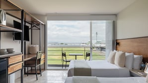 Quarto de hotel com cama, escrivaninha e cadeira. Há uma janela com vista para uma área gramada e edifícios.