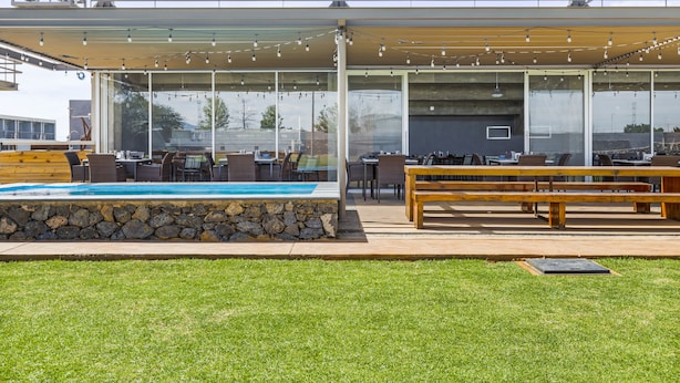 Área de piscina ao ar livre com paredes de pedra, bancos e mesas de madeira sob um alpendre coberto com luzes de corda.