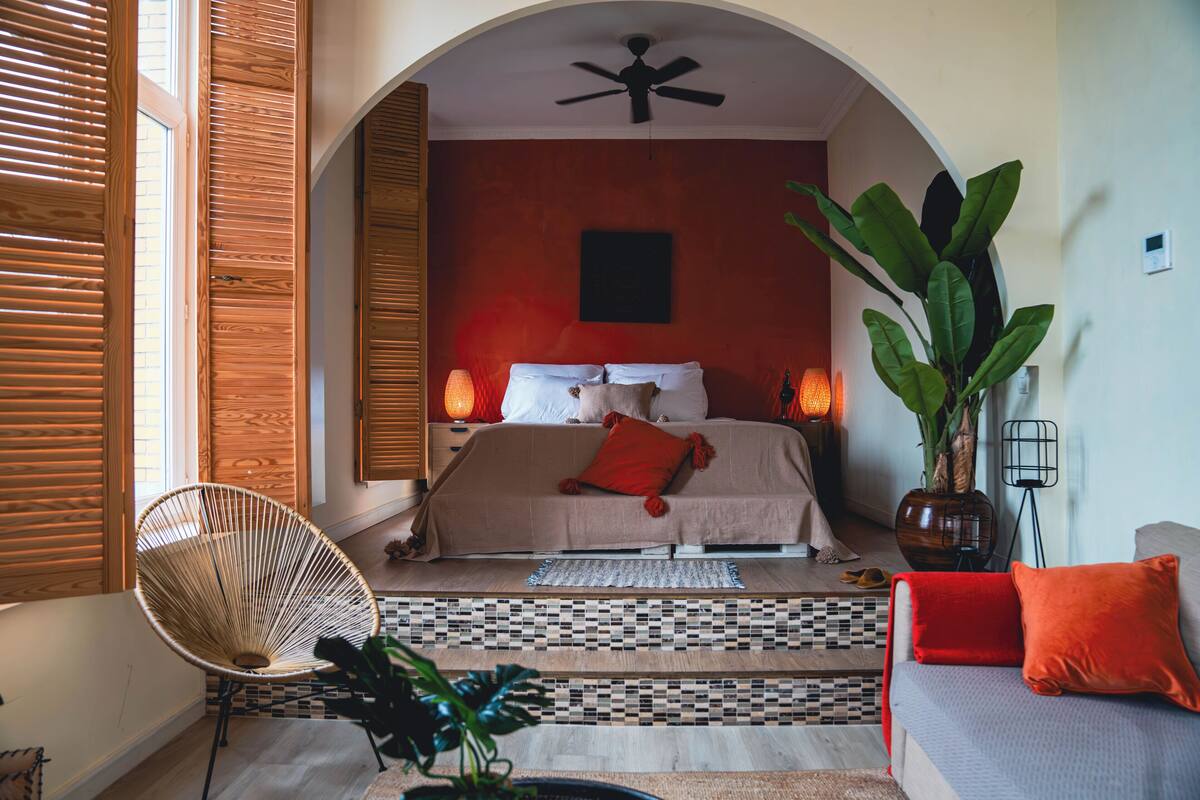 Quarto com cama, poltrona, sofá, ventoinha no teto e uma planta grande.